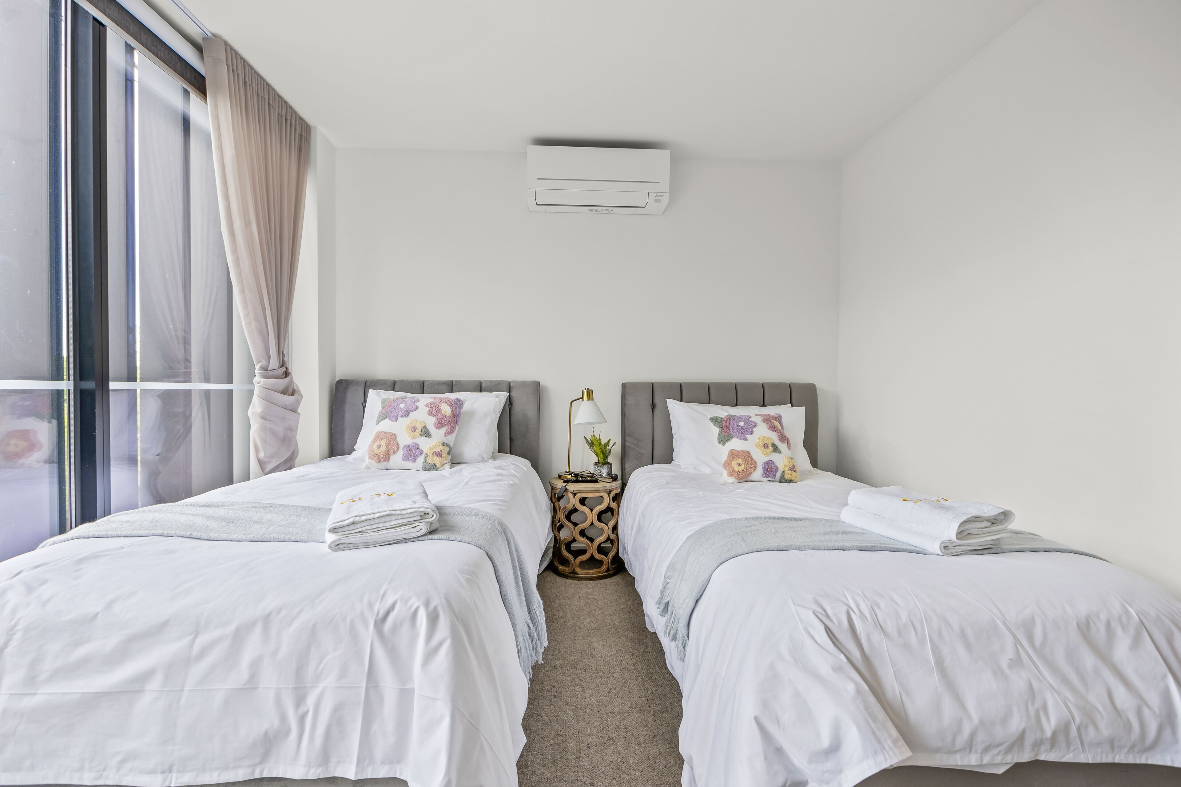 Quarto de hotel com duas camas, uma janela com cortinas, um ar condicionado, uma mesinha de cabeceira com luminária e um pequeno vaso de planta.
