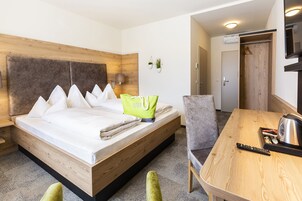 Quarto de hotel com cabeceira de madeira, cama com roupa de cama branca, uma mesinha com chaleira, uma cadeira e um controle remoto.