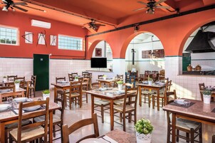 Um restaurante com mesas e cadeiras de madeira, uma televisão presa à parede e uma cozinha visível através de um arco.