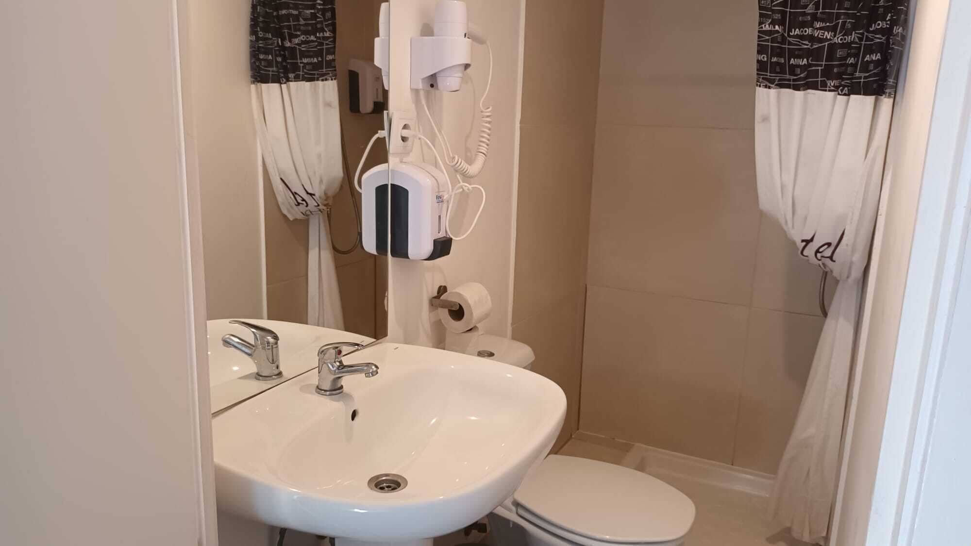 Casa de banho com lavatório branco, duas torneiras, sanita, secador de cabelo e suporte para toalhas.