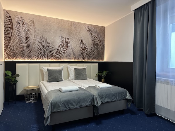 Quarto de hotel moderno com uma cama grande, duas mesas de cabeceira, um painel de luz fixado na parede e vista para o exterior através de uma janela com cortinas.