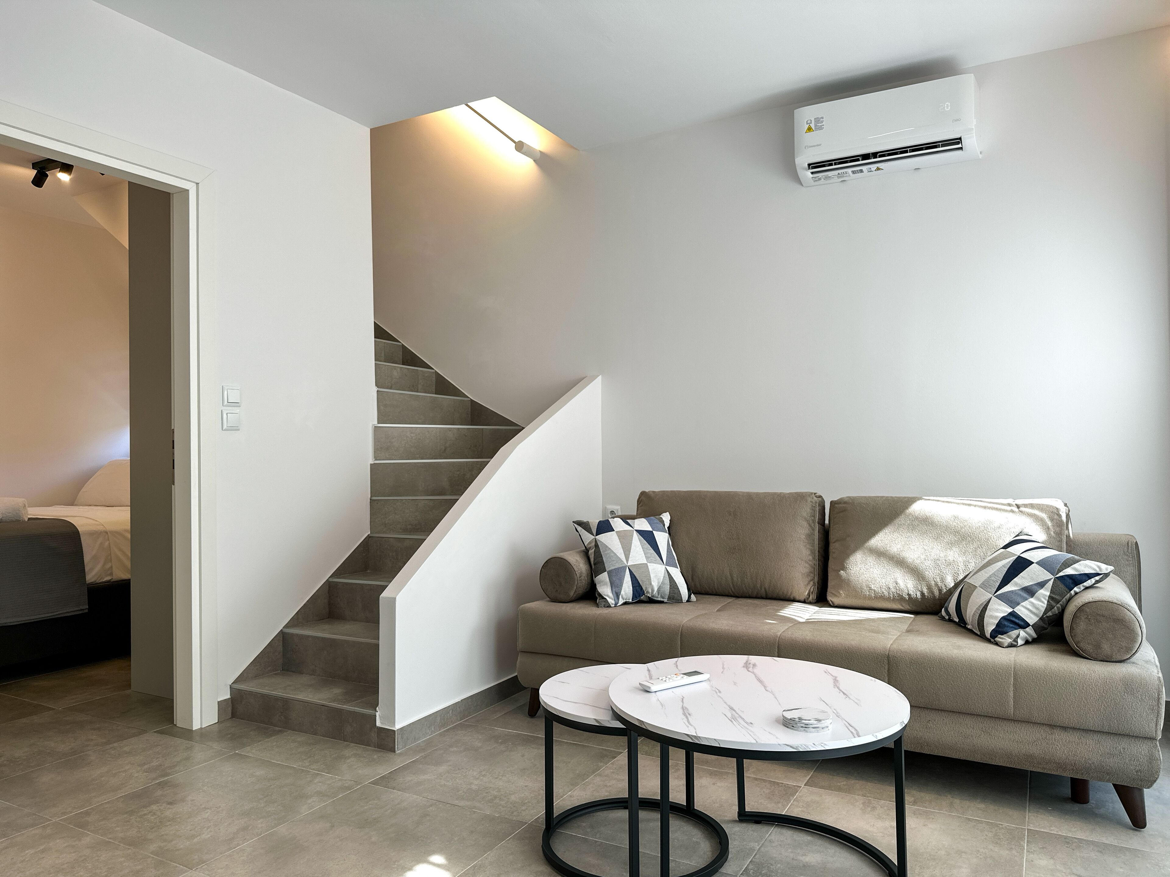Uma sala de estar moderna com escada, um sofá bege, duas mesas redondas e um aparelho de ar condicionado.