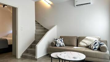 Uma sala de estar moderna com escada, um sofá bege, duas mesas redondas e um aparelho de ar condicionado.