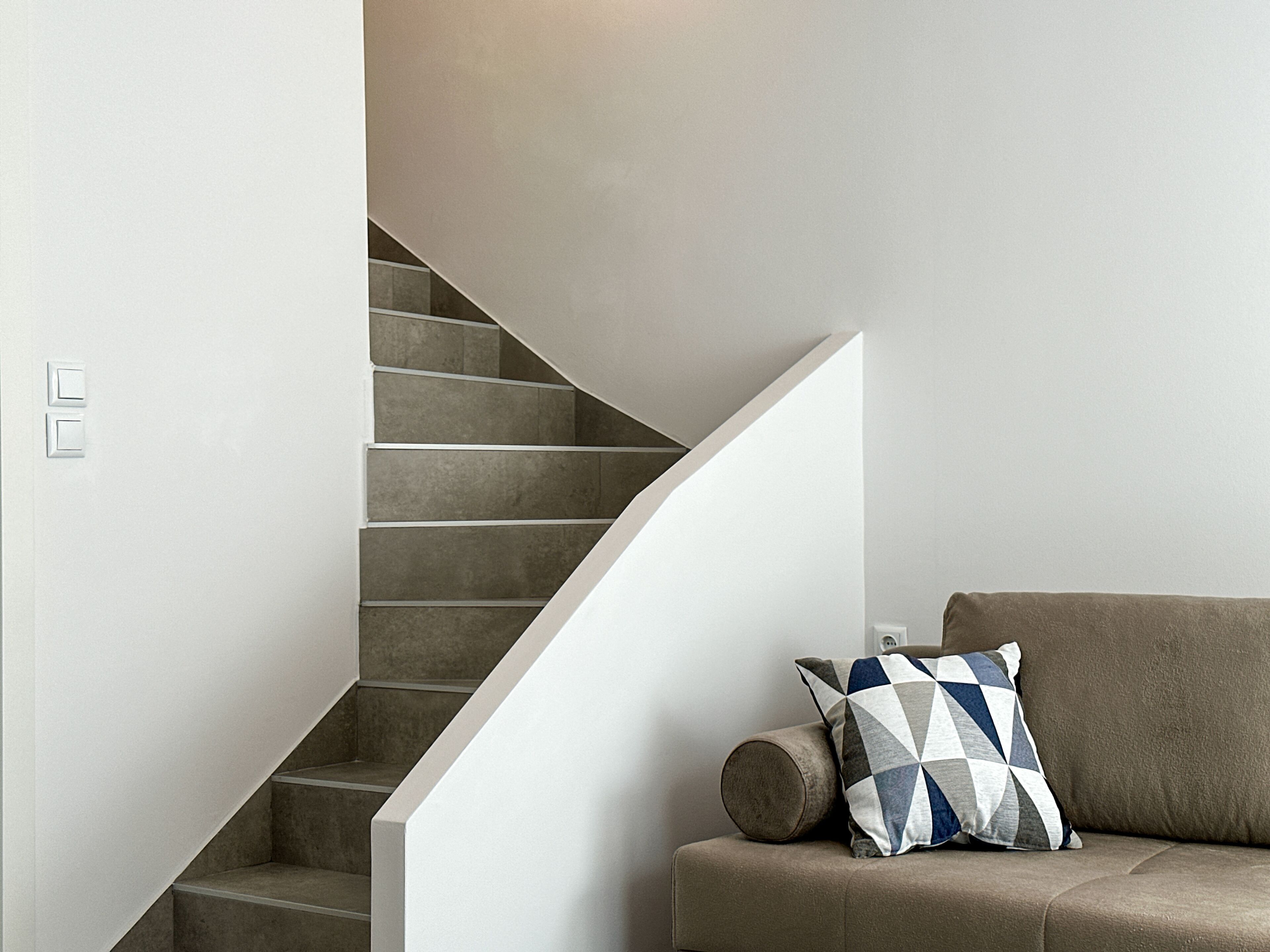 Uma escadaria com design moderno, um sofá com almofadas estampadas e um interruptor de parede.