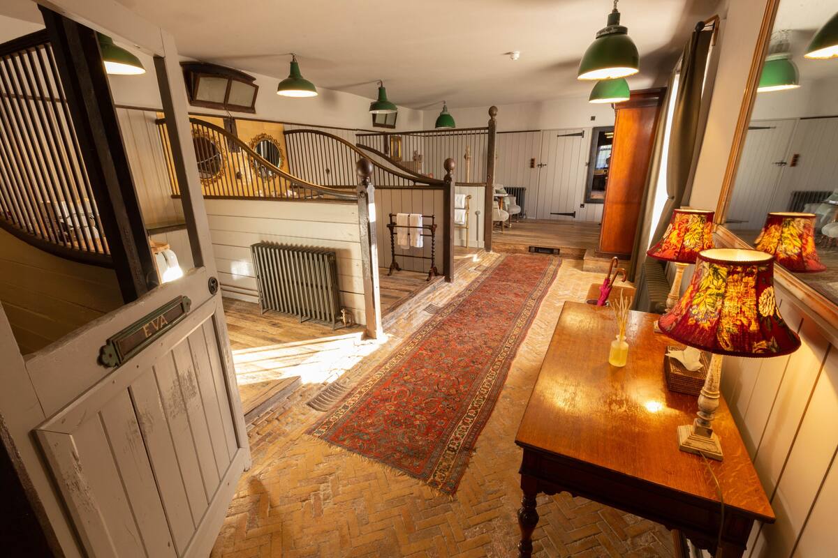 Um corredor com piso de madeira, um tapete vermelho estampado, uma mesa de madeira com luminárias e uma escada com corrimão.