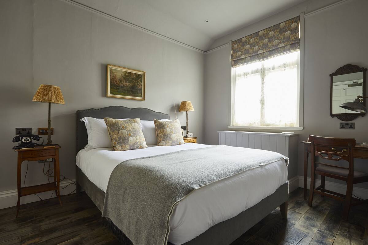 Quarto bem arrumado com uma cama grande, duas mesas de cabeceira, uma cadeira, um quadro na parede e uma janela com cortinas.