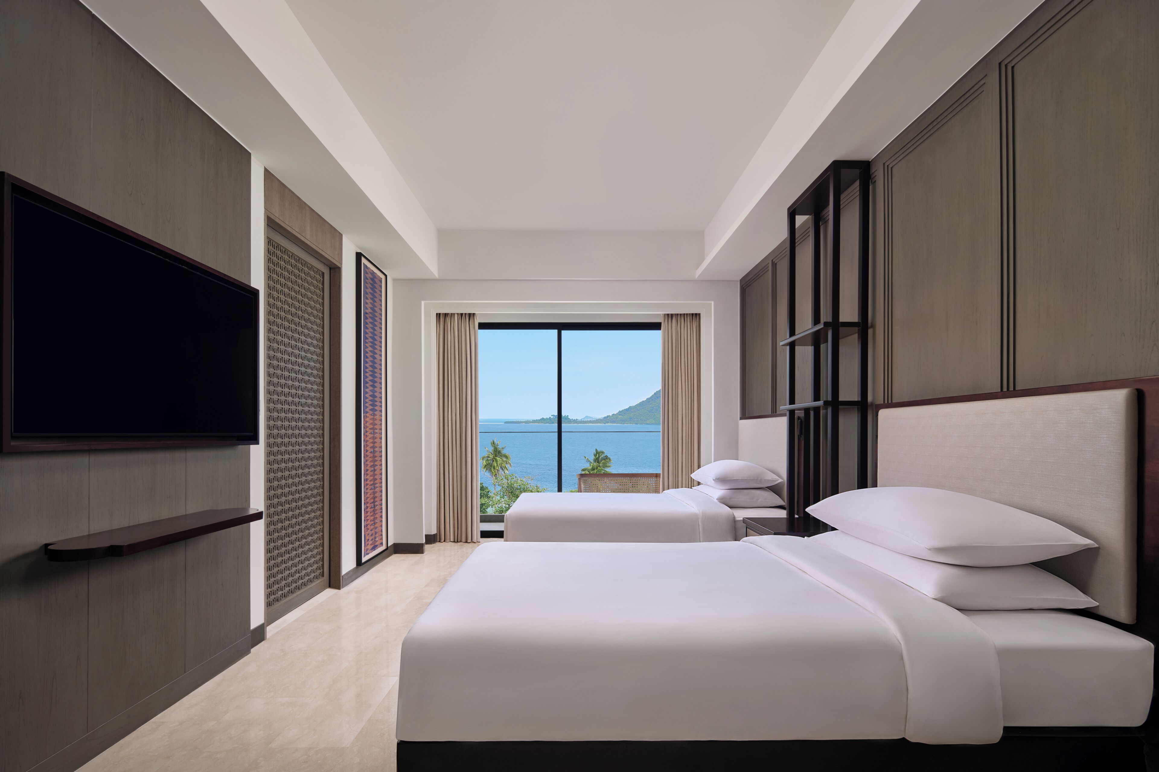 Quarto de hotel com uma televisão grande, duas camas, vista para o mar e varanda.
