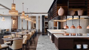 Interior moderno de restaurante com várias mesas e cadeiras, área de bar central com panelas de cobre e luminárias pendentes.