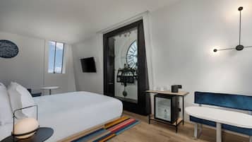 Quarto de hotel moderno com uma cama grande, uma pequena escrivaninha, um sofá azul, uma televisão, um relógio e uma janela com vista para uma torre do relógio.