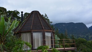Uma pequena cabana de madeira com telhado inclinado, cercada por vegetação exuberante e montanhas ao fundo.