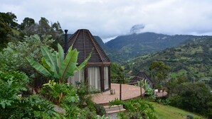 Uma casa com telhado inclinado, rodeada por vegetação exuberante e montanhas ao fundo.
