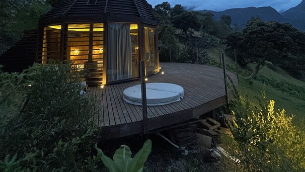 Uma cabana de madeira com terraço circular e banheira de hidromassagem, rodeada por vegetação exuberante e colinas.