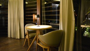 Um quarto aconchegante com uma mesa redonda e duas cadeiras, uma luminária e vista para o exterior através de amplas janelas com cortinas.