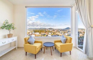 Quarto com duas poltronas amarelas, uma mesa redonda pequena, um vaso de planta e uma grande janela com vista para a cidade.