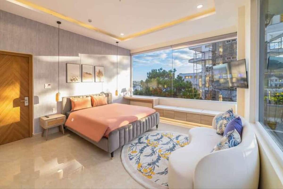 Um quarto moderno com uma cama grande, uma mesa de cabeceira, uma luminária de apoio, uma janela com vista para edifícios e um espaço de estar circular com um tapete estampado.