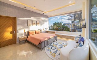Um quarto moderno com uma cama grande, uma mesa de cabeceira, uma luminária de apoio, uma janela com vista para edifícios e um espaço de estar circular com um tapete estampado.