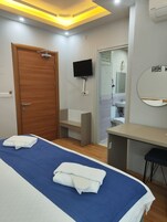 Quarto de hotel com cama, escrivaninha, televisão na parede e banheiro visível através de uma porta aberta.