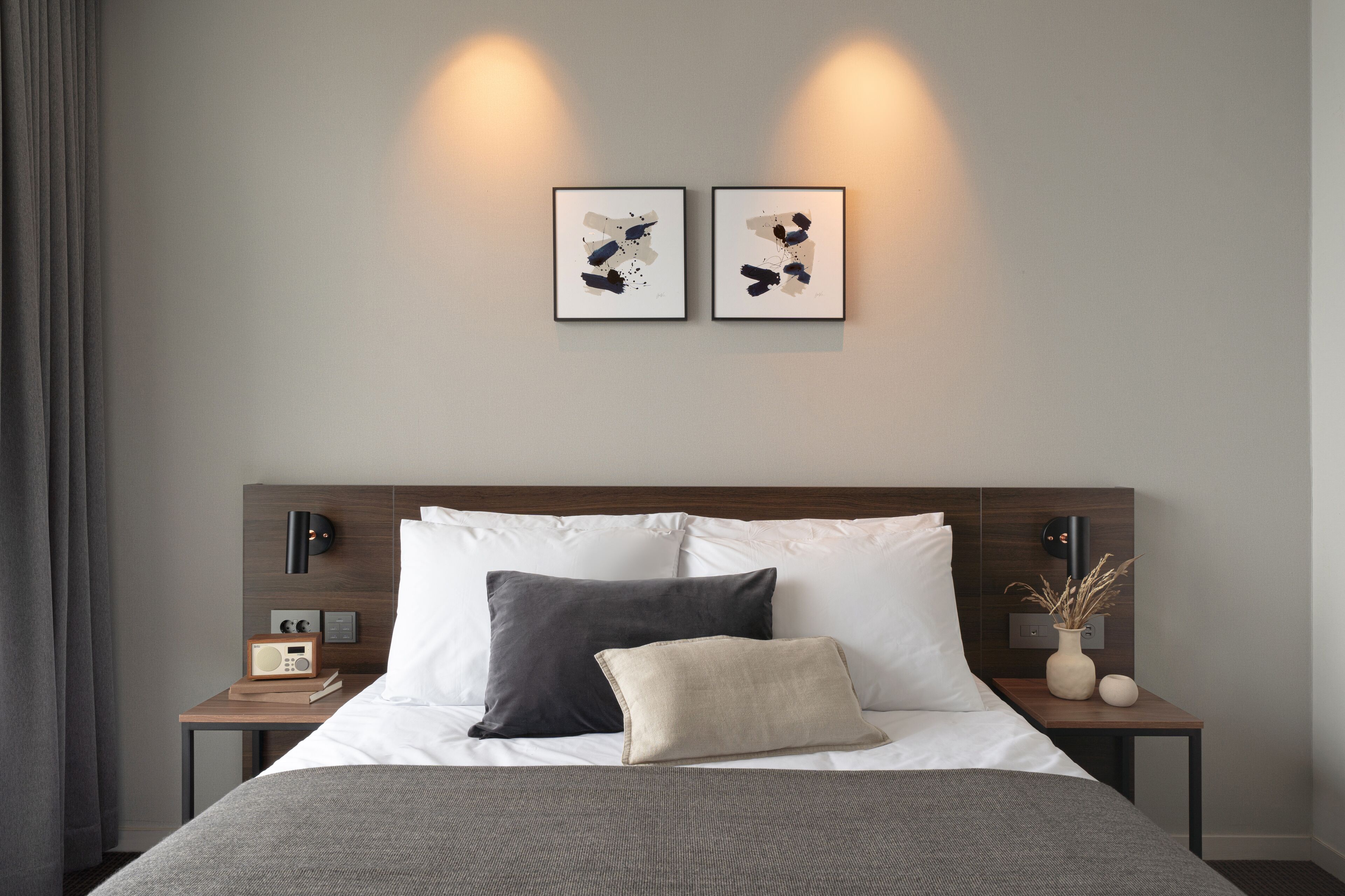 Quarto de hotel bem arrumado, com uma cama, duas mesas de cabeceira, um rádio e luminárias fixadas na parede.