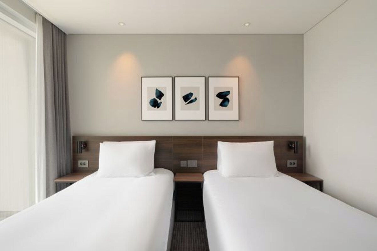 Quarto de hotel com duas camas de solteiro, cabeceira de madeira, uma obra de arte na parede e uma janela com cortinas.