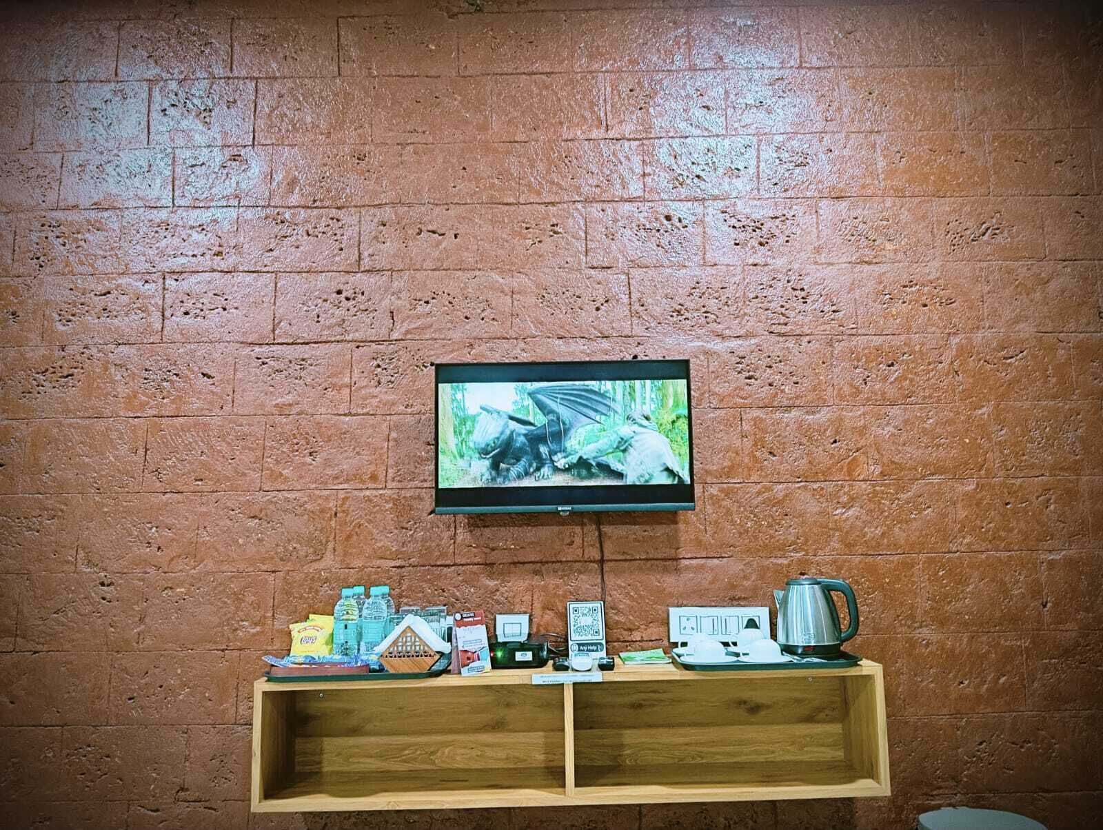 Uma televisão fixada numa parede de tijolos, uma prateleira de madeira com vários objetos, e uma chaleira.