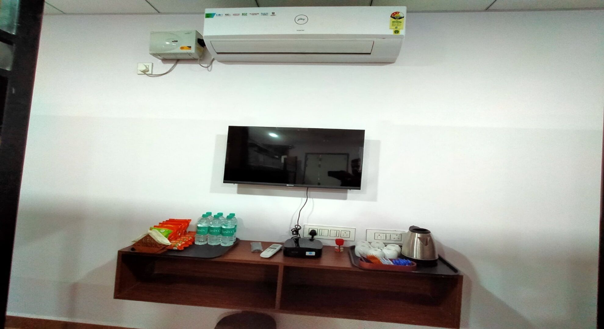 Quarto com televisão presa na parede, escrivaninha de madeira com um comando, garrafas de água, chaleira e régua de tomadas.