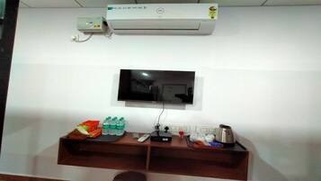 Quarto com televisão presa na parede, escrivaninha de madeira com um comando, garrafas de água, chaleira e régua de tomadas.
