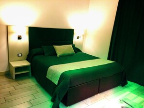 Uma cama bem arrumada com colcha e almofadas verdes, uma mesa de cabeceira e duas luminárias fixadas na parede.