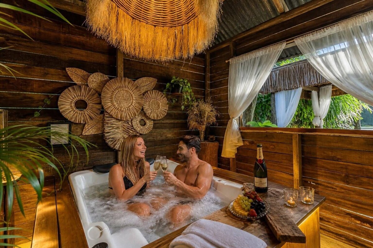 Um casal em quarto de cabana rústica com hidromassagem, rodeado por decorações de parede em tear e uma garrafa de champanhe.