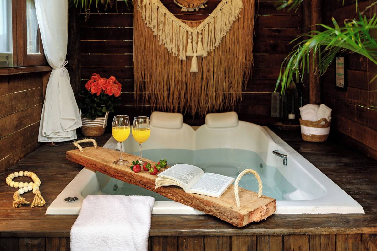 Uma cabana de madeira com hidromassagem, um tabuleiro de madeira com um livro, copos de suco de laranja e um buquê de flores vermelhas.