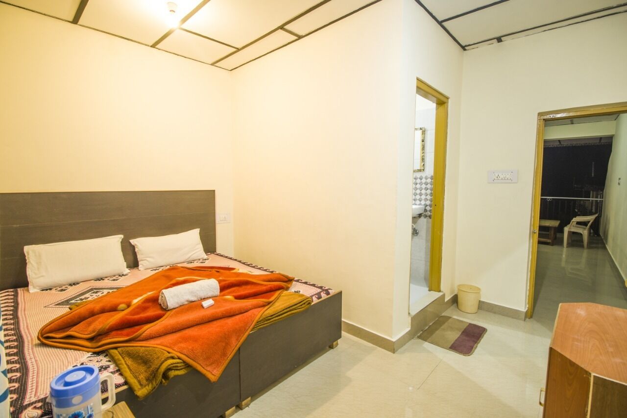 Quarto de hotel com uma cama, uma mesinha, uma cadeira e uma porta que dá para outro cômodo.