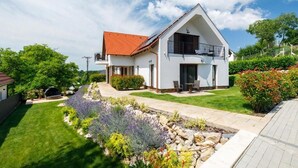 Uma casa moderna de dois andares com telhado inclinado, varanda e um jardim bem cuidado com diversas plantas e um muro de arrimo de pedra.
