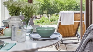 Uma área de refeições com uma mesa posta para dois, uma cadeira de vime e vista para uma paisagem verde através de grandes janelas.
