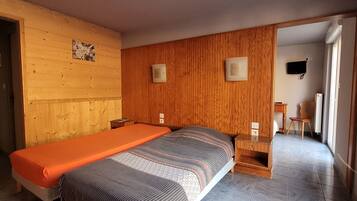 Quarto com cama, paredes e porta de madeira, uma mesinha, uma cadeira e televisão fixada na parede.