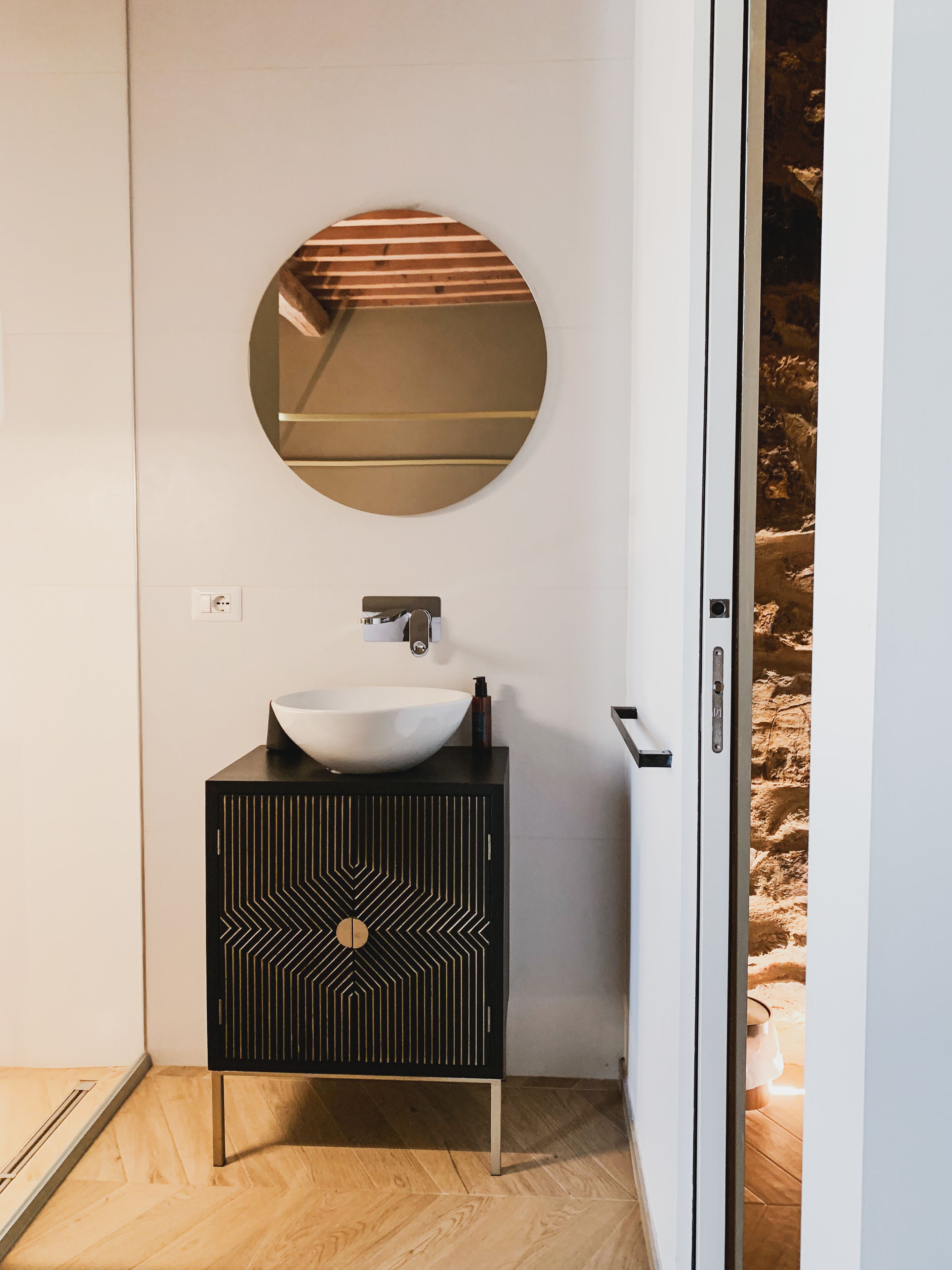Uma casa de banho com uma pia branca, um armário preto com padrões geométricos, um espelho redondo e um chão de madeira.