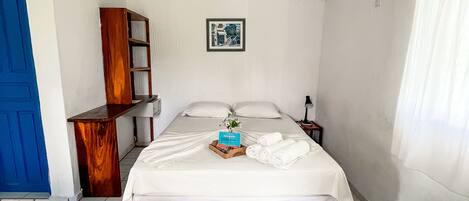 Quarto com cama, um pequeno armário de madeira, um quadro pendurado na parede e uma porta azul.