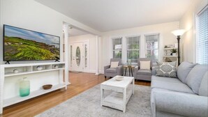 Uma sala de estar com televisão de tela plana, sof ás cinzas, uma mesa de centro e um rack para aparelhos branco.