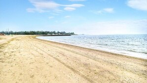 Uma praia arenosa com ondas suaves, céu claro e uma linha de costa distante com árvores e edifícios.