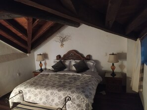 Quarto com teto de madeira, cama com roupa de cama estampada, duas mesas de cabeceira com abajures e um adorno fixado na parede.