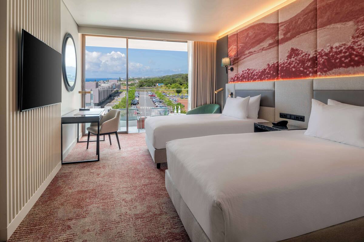 Quarto de hotel com duas camas, uma escrivaninha, televisão e uma janela grande com vista para a cidade.