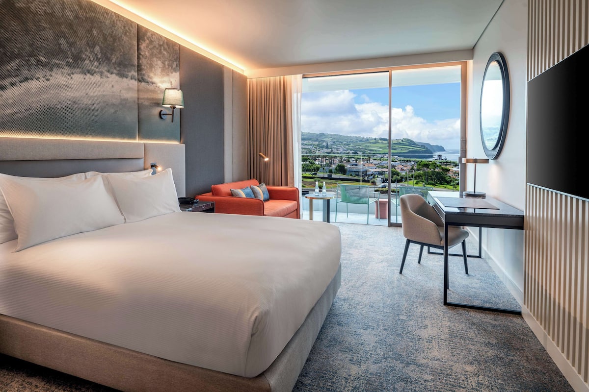 Quarto de hotel com uma cama grande, uma escrivaninha, uma cadeira, uma televisão, uma janela ampla com vista para a cidade e uma luminária na parede.