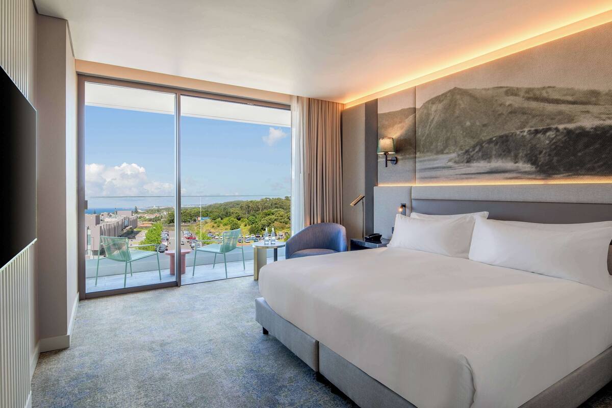 Quarto de hotel com uma cama grande, uma escrivaninha, uma cadeira, televisão e varanda com vista para a cidade e áreas verdes.
