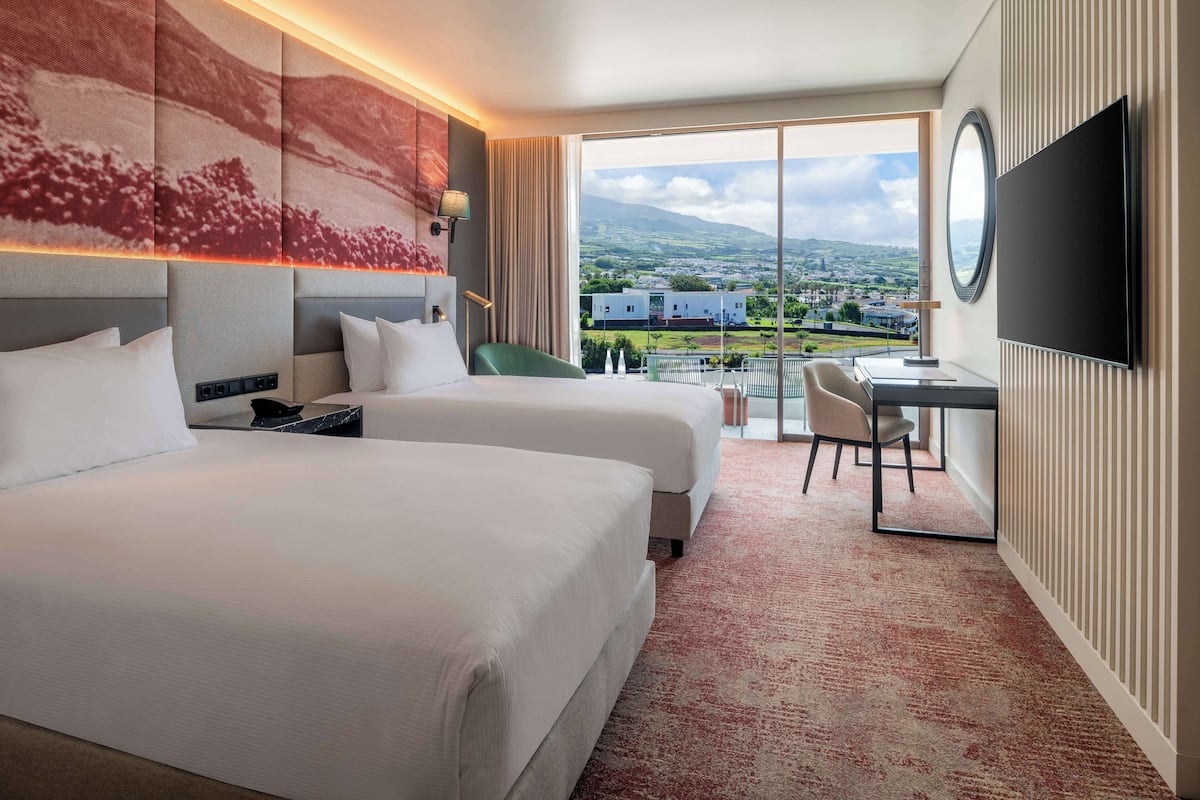 Quarto de hotel com duas camas, uma escrivaninha com cadeira, televisão e uma janela grande com vista para a cidade.