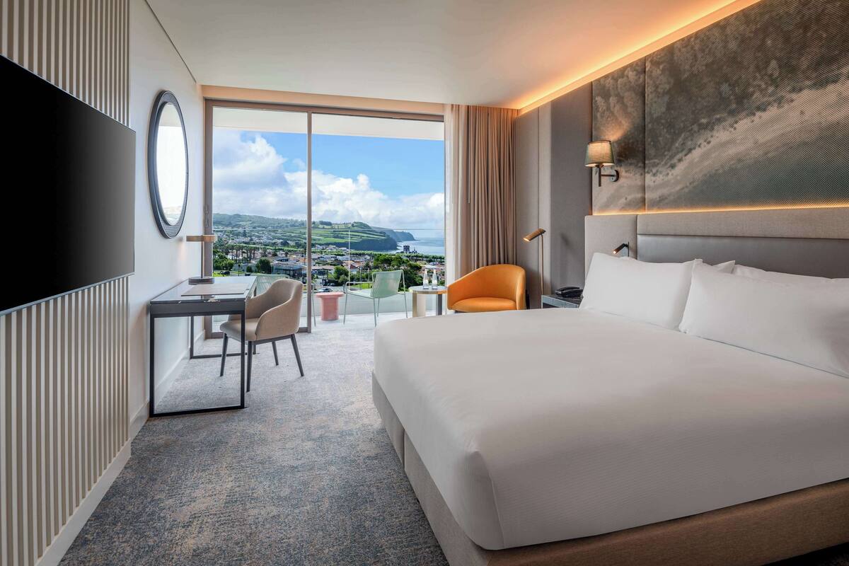 Quarto de hotel com uma cama grande, uma escrivaninha com cadeira, televisão e vista para a cidade e colinas.