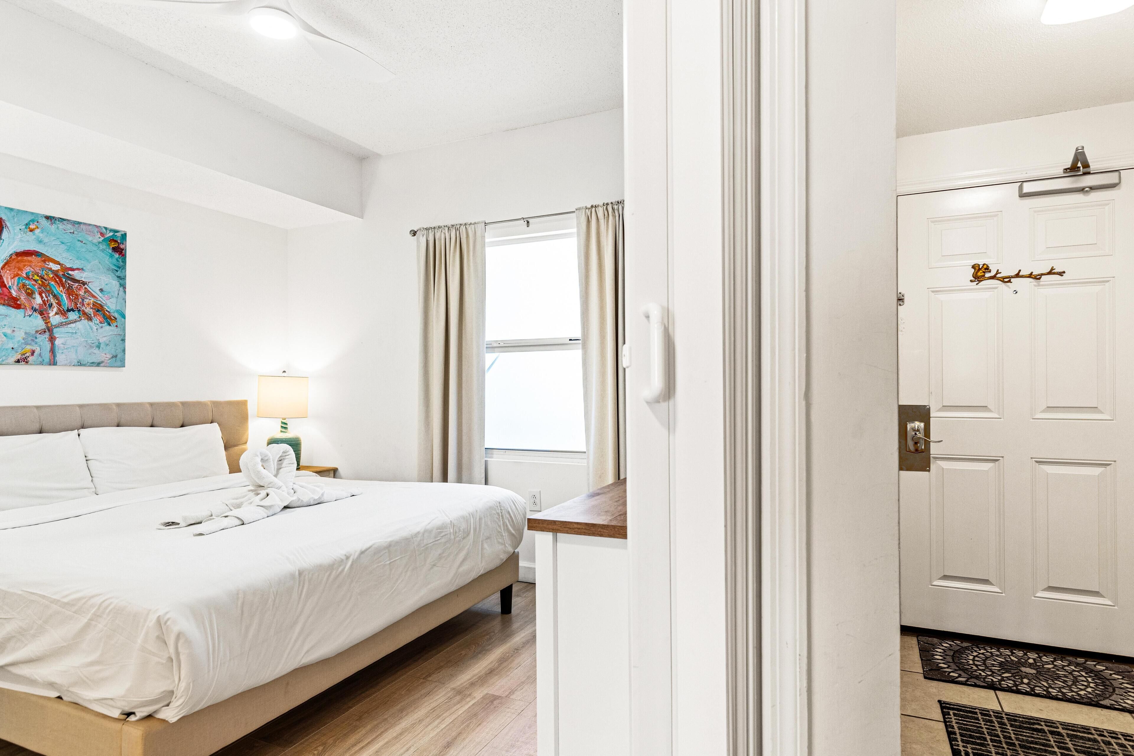 Quarto de hotel com cama, janela com cortinas, porta com chave na fechadura e um quadro na parede.