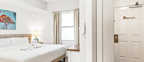 Quarto de hotel com cama, janela com cortinas, porta com chave na fechadura e um quadro na parede.