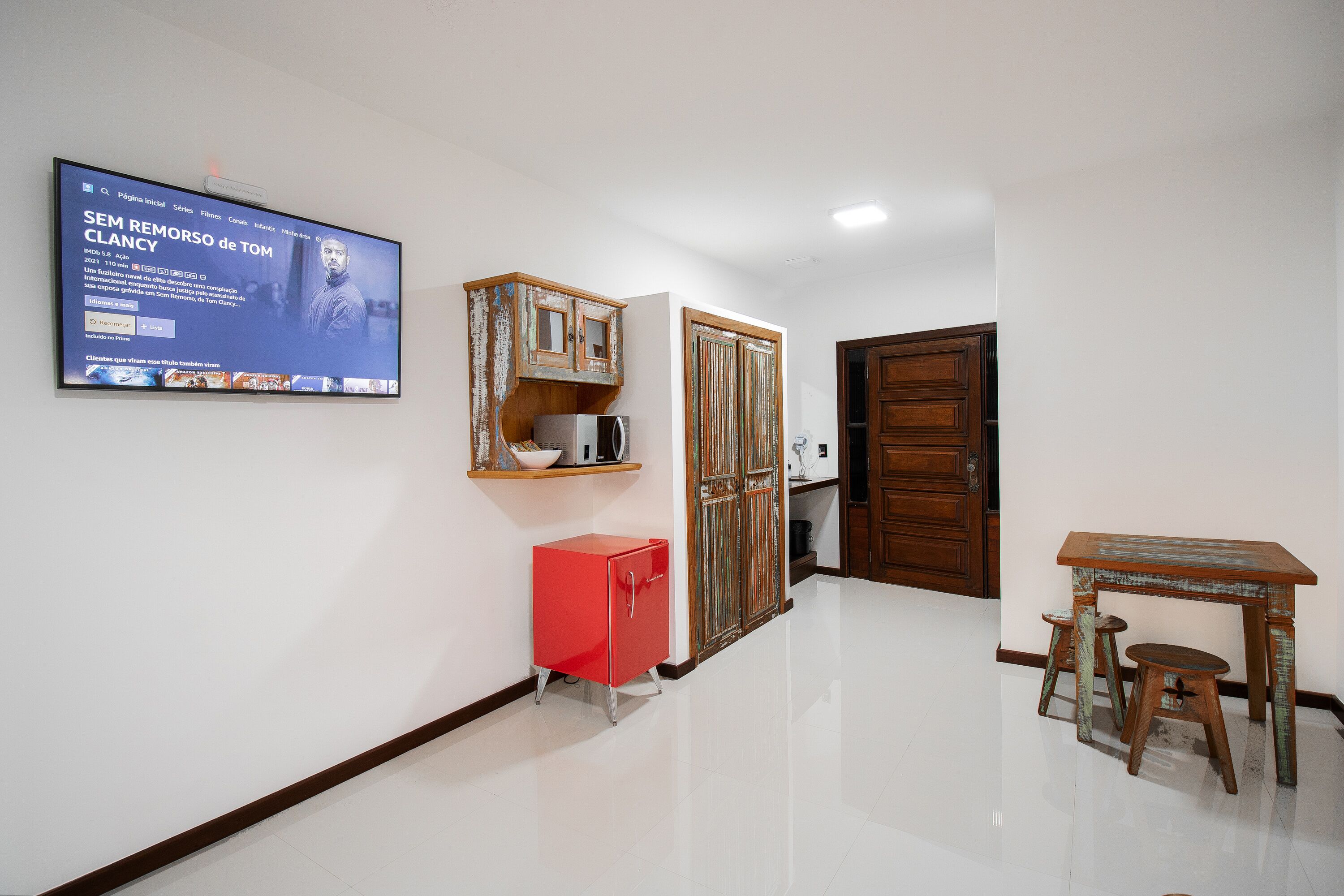 Um corredor com uma televisão exibindo um canal de notícias, um armário vermelho, um aparador de madeira com espelho e uma mesa de madeira com bancos.