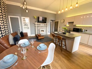 Uma cozinha moderna com uma ilha central, uma sala de jantar com mesa de madeira e cadeiras brancas, e uma sala de estar com sofá de couro marrom.