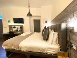 Quarto de hotel moderno com uma cama grande, uma escrivaninha com televisão de tela plana, uma kitchenette e uma parede de destaque em madeira.