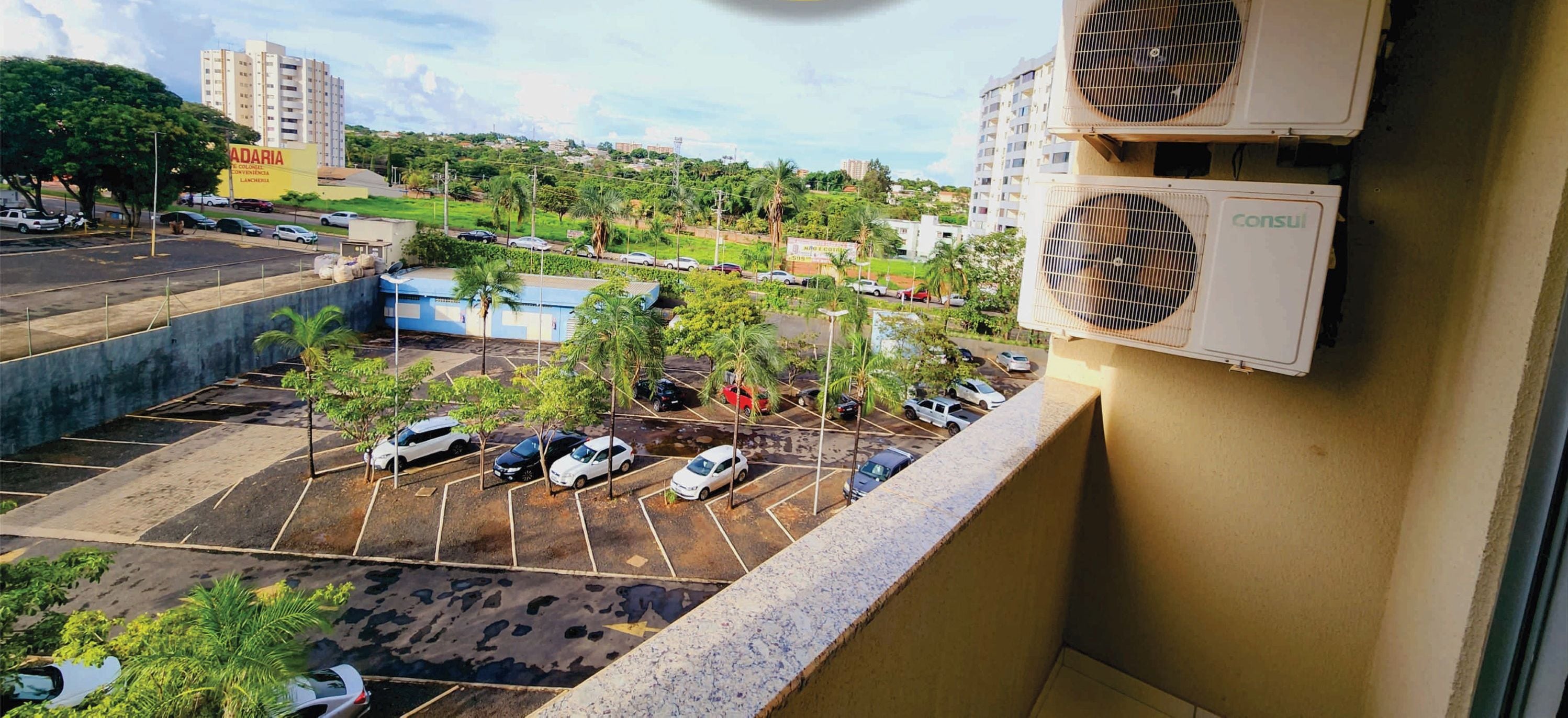 Vista de uma varanda com vista para um estacionamento com vários carros, palmeiras e edifícios ao fundo.