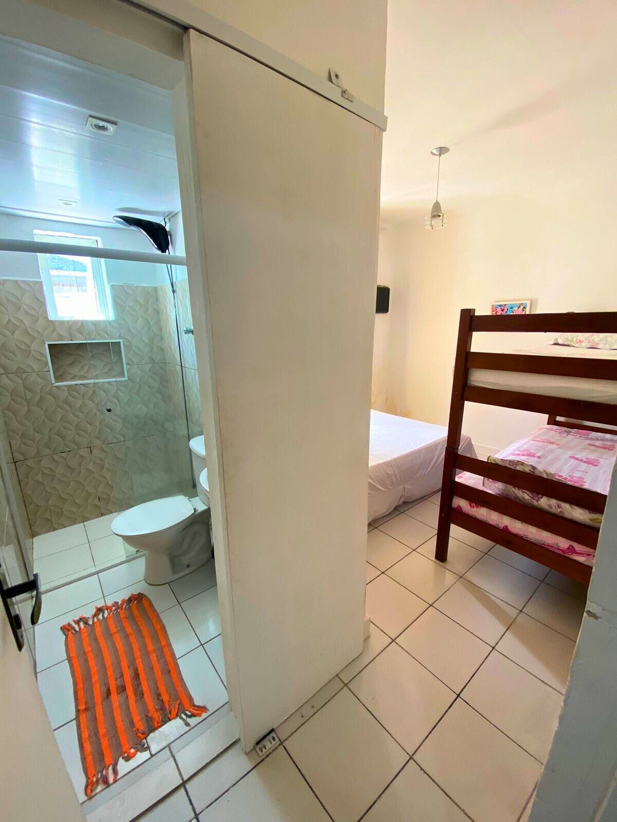 Quarto pequeno e limpo, com chuveiro, sanitário e uma cama.